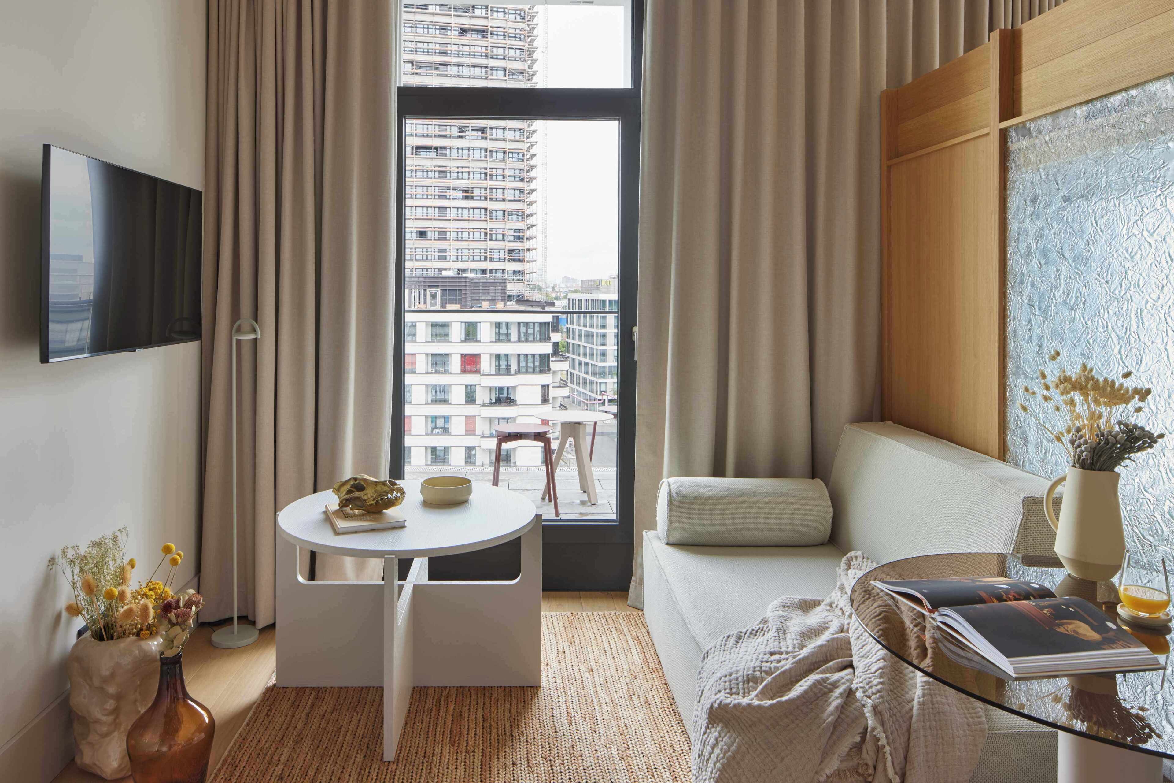 Quarto de hotel moderno com uma grande janela, televisão de tela plana, mesa redonda pequena, sofá e mesa de centro de vidro.