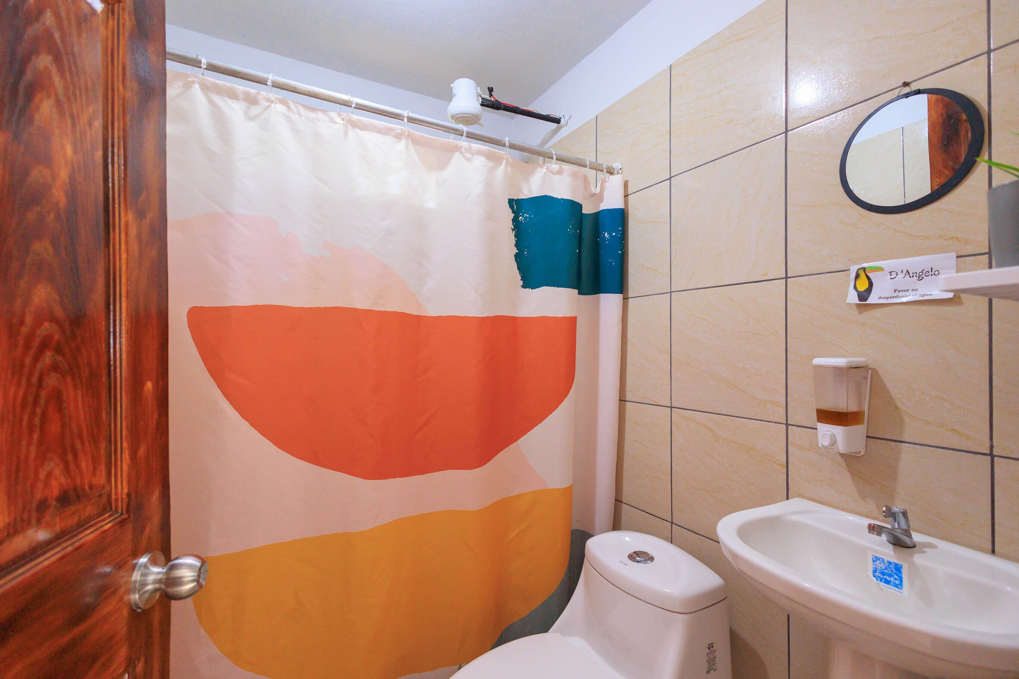 Um banheiro com porta de madeira, cortina de chuveiro com padrão geométrico, vaso sanitário, pia com espelho acima e dispensador de sabonete na parede.