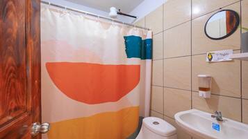 Um banheiro com porta de madeira, cortina de chuveiro com padrão geométrico, vaso sanitário, pia com espelho acima e dispensador de sabonete na parede.
