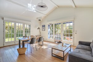 Uma sala de estar ampla com piso de madeira, um sofá cinza, uma mesa de jantar com cadeiras e grandes janelas com vista para o verde.
