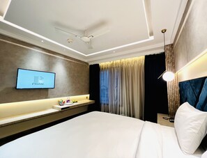 Quarto de hotel moderno com televisão de tela plana exibindo o logotipo 'PRIME', um ventilador de teto, mesa de cabeceira com luminária e uma cama grande com lençóis brancos.