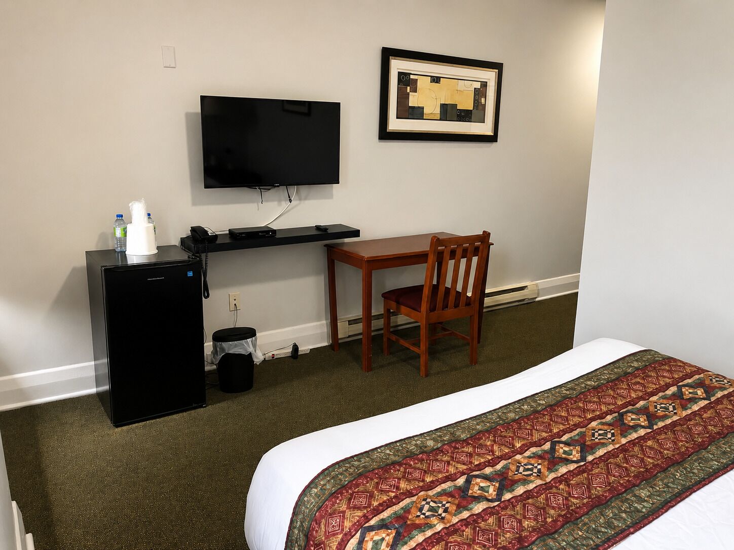 Quarto de hotel com cama, escrivaninha e cadeira, televisão e quadro na parede.
