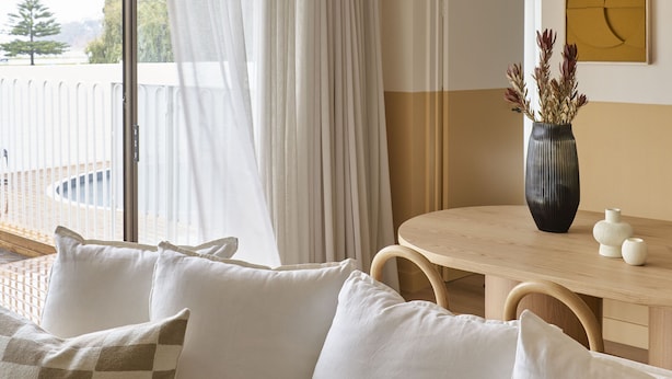 Quarto de hotel com sofá, mesa de jantar, vaso com flores secas e vista para o exterior.