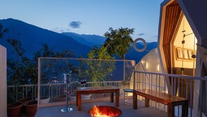 Um terraço moderno ao ar livre com lareira, espaço para sentar e guarda-corpo de vidro, com vista para uma paisagem montanhosa.