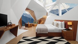 Quarto de hotel moderno com uma cama grande, uma escrivaninha e uma televisão. O quarto tem vista para as montanhas e um claraboia.