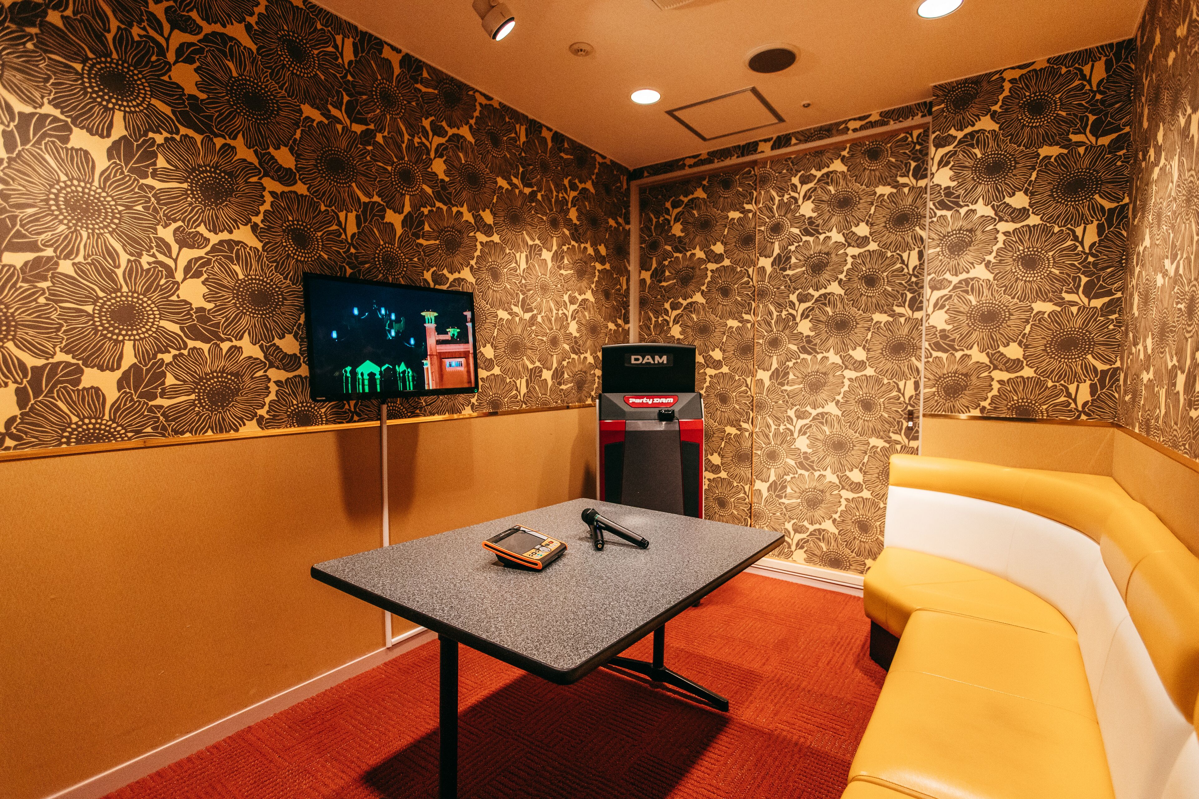 Quarto com parede estampada, televisão de tela plana, máquina DAM, pequena mesa com telefone e controle remoto, e sofá amarelo.