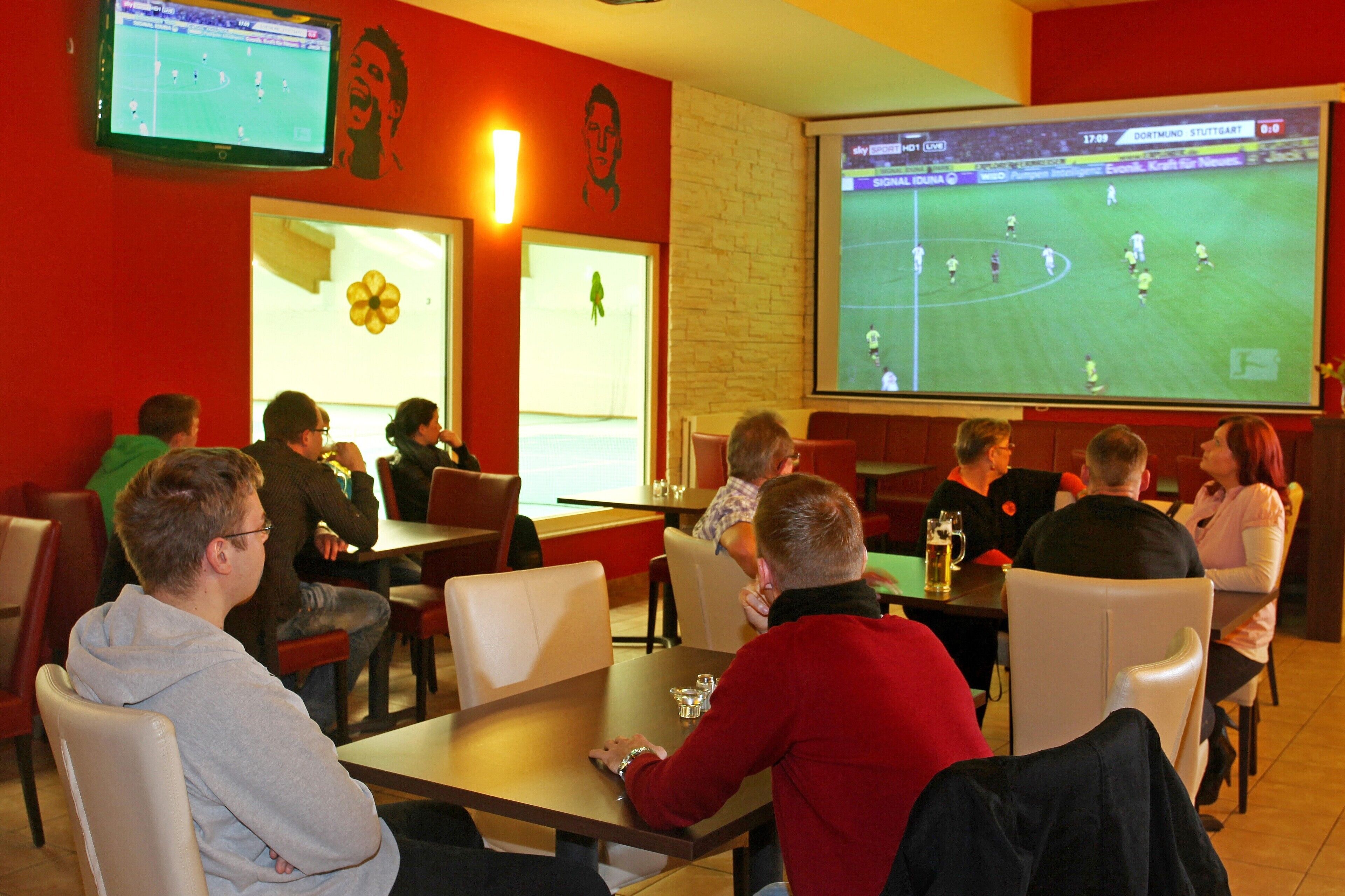 Um grupo de pessoas sentadas em mesas num restaurante, com um ecrã grande a exibir um jogo de futebol.