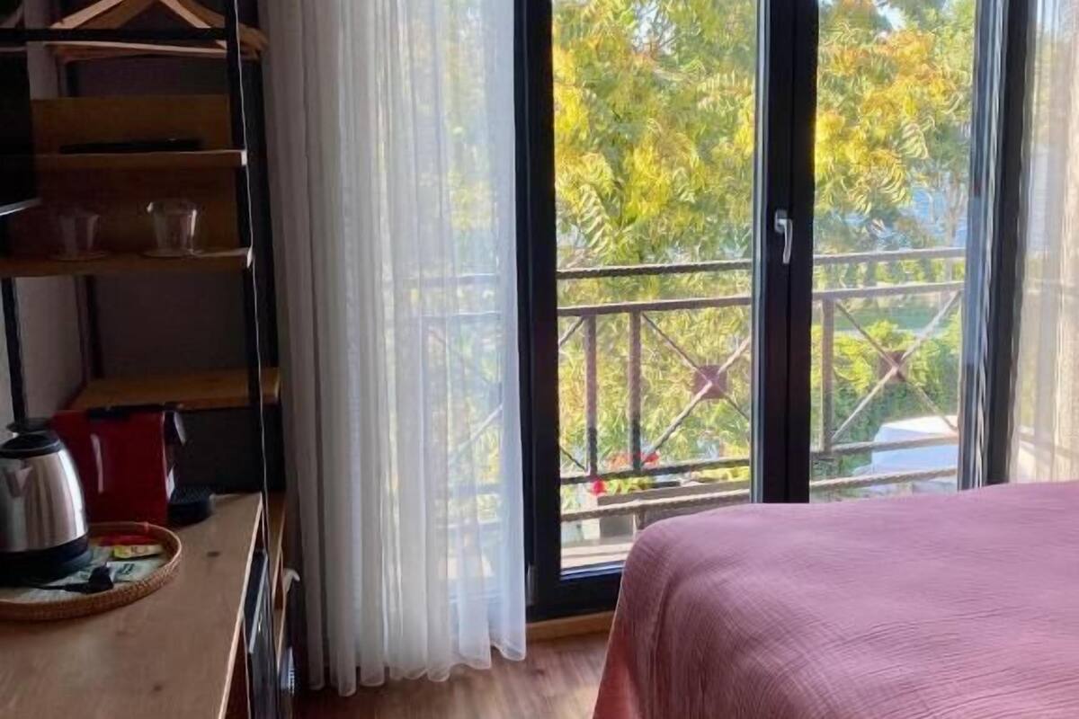 Quarto de hotel com cama, uma escrivaninha de madeira com chaleira, uma prateleira com cabides e varanda com árvores.