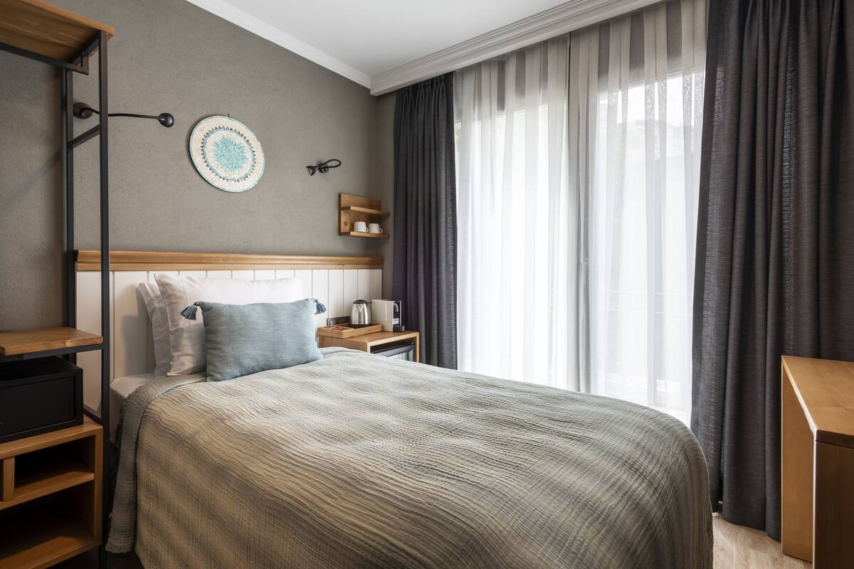 Quarto de hotel bem arrumado, com uma cama grande, mesas de cabeceira, prateleira na parede e uma janela com cortinas.