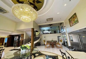 Um hall amplo com um lustre imponente, uma mesa de vidro central com uma planta e uma escadaria para um andar superior.