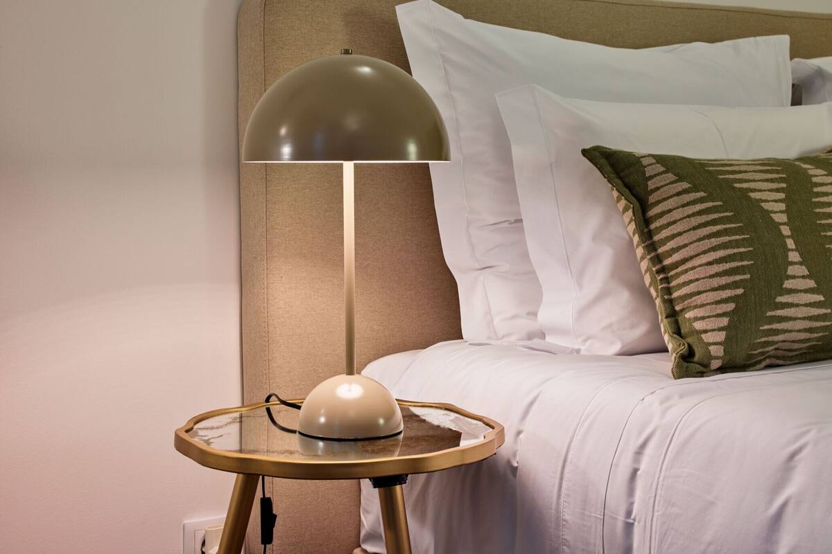 Uma cama com roupa de cama branca e um travesseiro estampado em verde, uma mesa redonda com abajur e um aplique na parede.