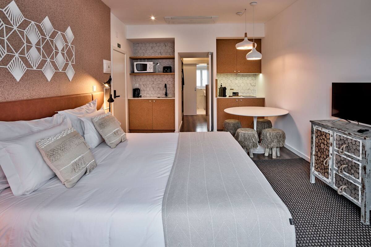 Quarto de hotel com cama, pequeno espaço de cozinha, área de refeições e televisão.