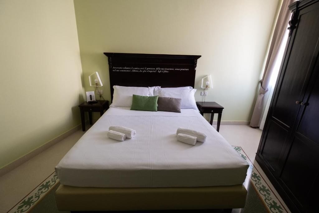 Cama bem arrumada com lençóis brancos e duas toalhas, uma cabeceira de madeira escura e duas luminárias de cabeceira em mesas de apoio.