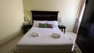 Cama bem arrumada com lençóis brancos e duas toalhas, uma cabeceira de madeira escura e duas luminárias de cabeceira em mesas de apoio.