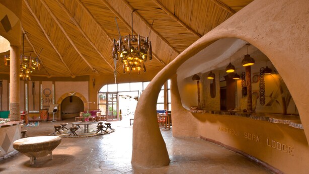 Um hall amplo com tetos altos, um pilar central de pedra e uma parede curva com o nome 'AMBOSELI SOPA LODGE'.
