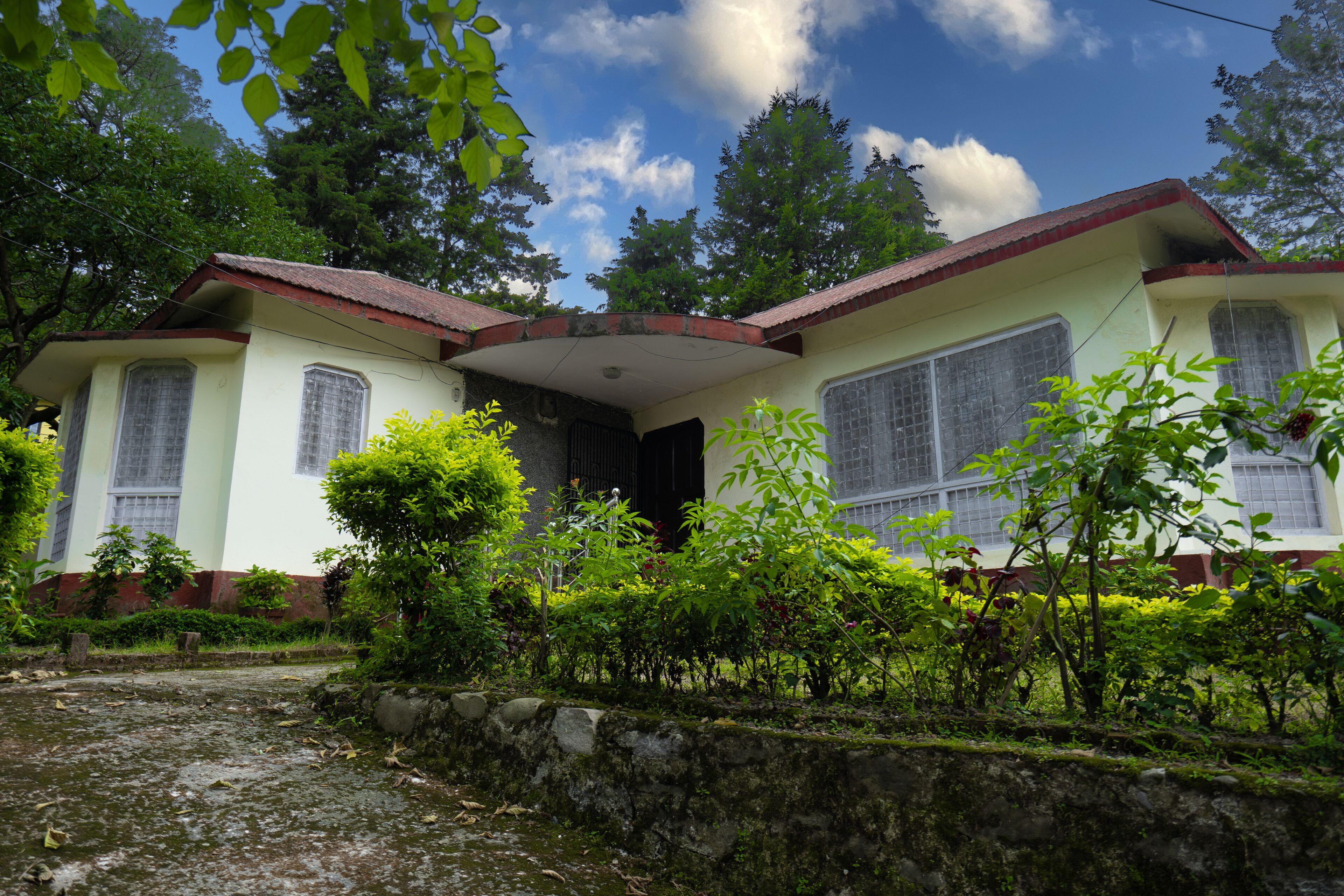 Uma casa de dois andares com telhado vermelho, cercada por vegetação exuberante e um pequeno riacho.