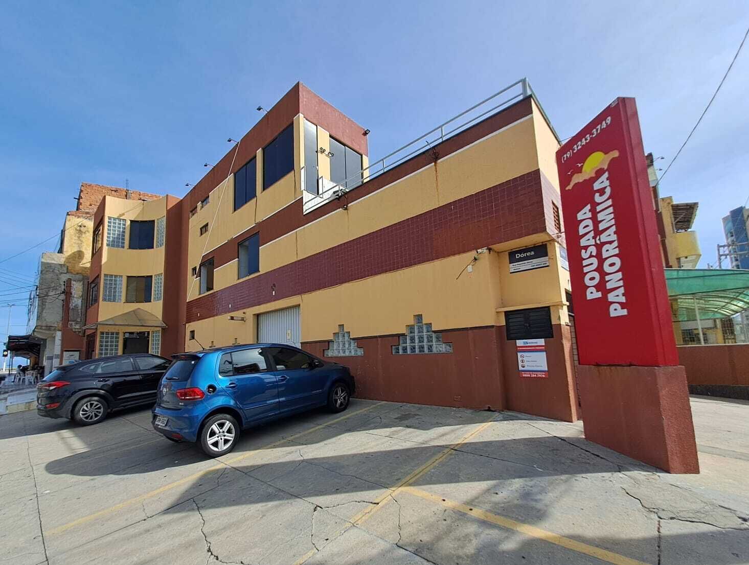 Um edifício com fachada vermelha e amarela, um carro azul estacionado na frente, e uma placa com a inscrição 'POUSADA PANORÁMICA'.