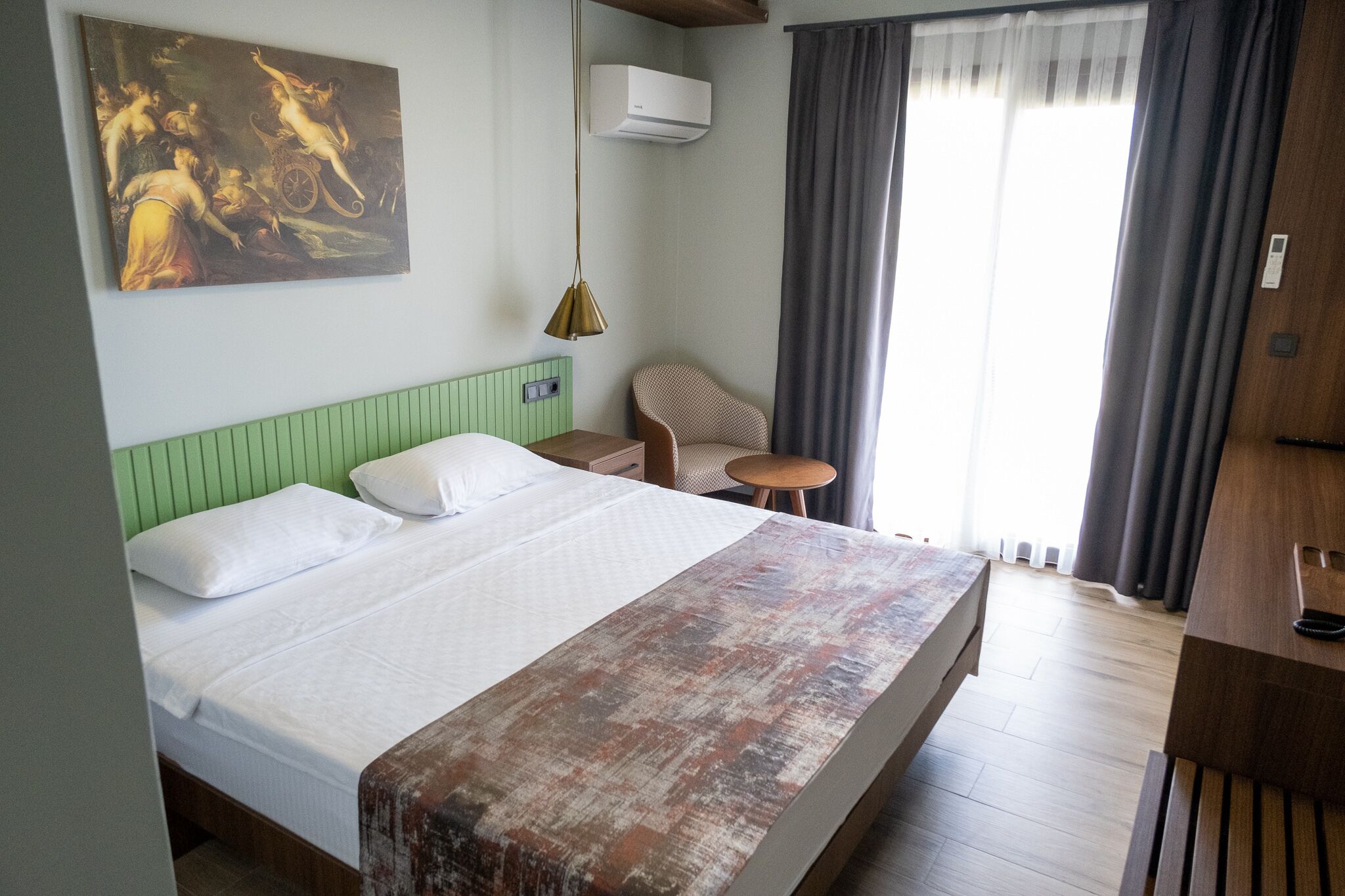 Quarto de hotel com cama grande, cabeceira verde, pintura na parede, mesa redonda pequena e janela com cortinas.