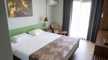 Quarto de hotel com cama grande, cabeceira verde, pintura na parede, mesa redonda pequena e janela com cortinas.