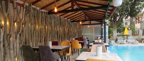 Restaurante com área à beira da piscina, apresentando vigas de madeira, plantas pendentes e cadeiras estofadas.