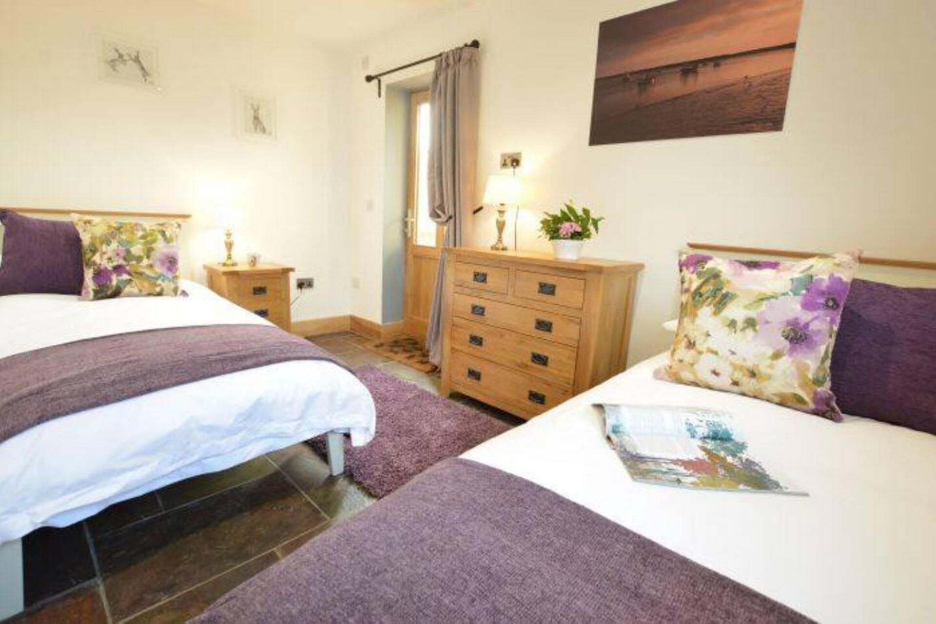 Quarto de hotel com duas camas, uma cômoda de madeira, uma mesinha de cabeceira e um quadro na parede.