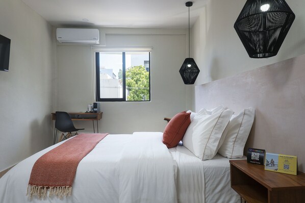 Quarto de hotel com cama, escrivaninha, cadeira, janela e luminária pendente preta.