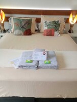 Cama bem arrumada com toalhas e um cartão, almofadas com estampas de folhas e cabeceira de madeira.