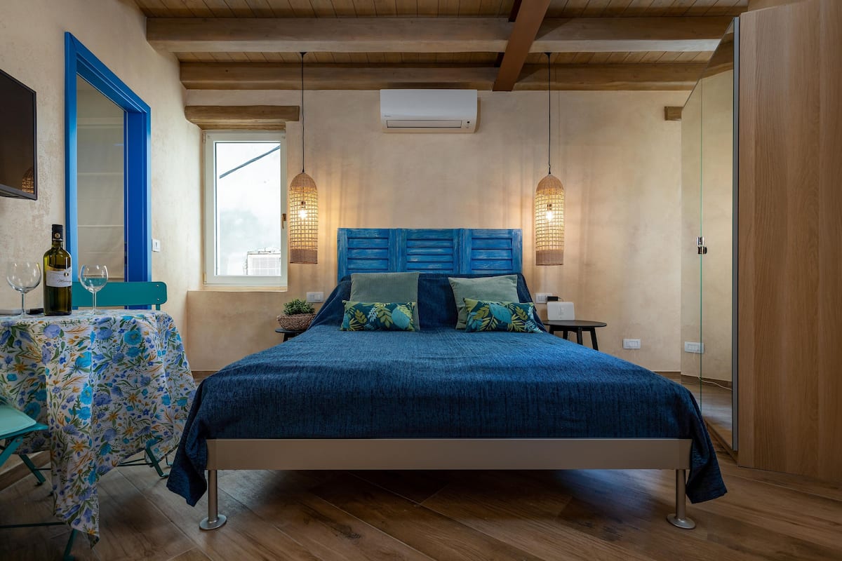 Quarto com teto de madeira, cama com cabeceira azul, duas luminárias pendentes e uma janela.