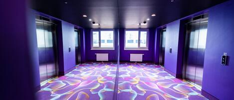 Um corredor com espelhos, elevadores e janelas, com um padrão de cores no chão.