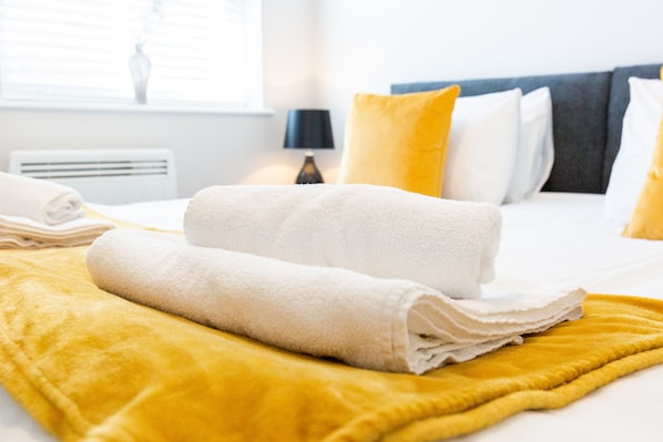 Cama bem arrumada com toalhas brancas e um cobertor amarelo, luminária de cabeceira e janela com persianas.
