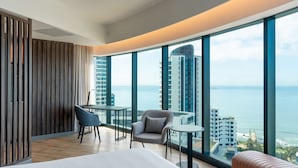 Quarto de hotel moderno com uma janela grande, uma cama, uma mesa pequena, duas cadeiras e vista para o oceano e edifícios.