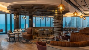 Um restaurante moderno com disposição circular de assentos, grandes janelas com vista para o mar e luminárias contemporâneas.