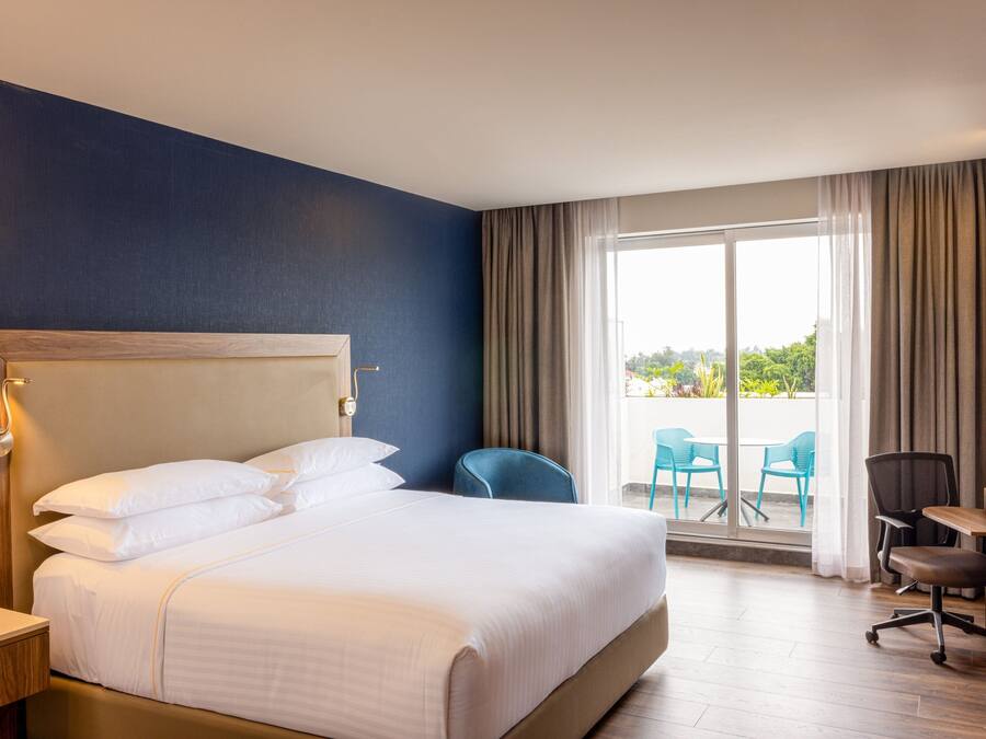 Quarto de hotel com uma cama grande, uma escrivaninha, uma cadeira e vista para o exterior através de uma porta de correr de vidro.
