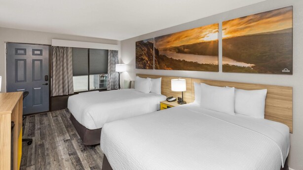 Quarto de hotel com duas camas, uma escrivaninha, uma cadeira, um abajur e uma porta.