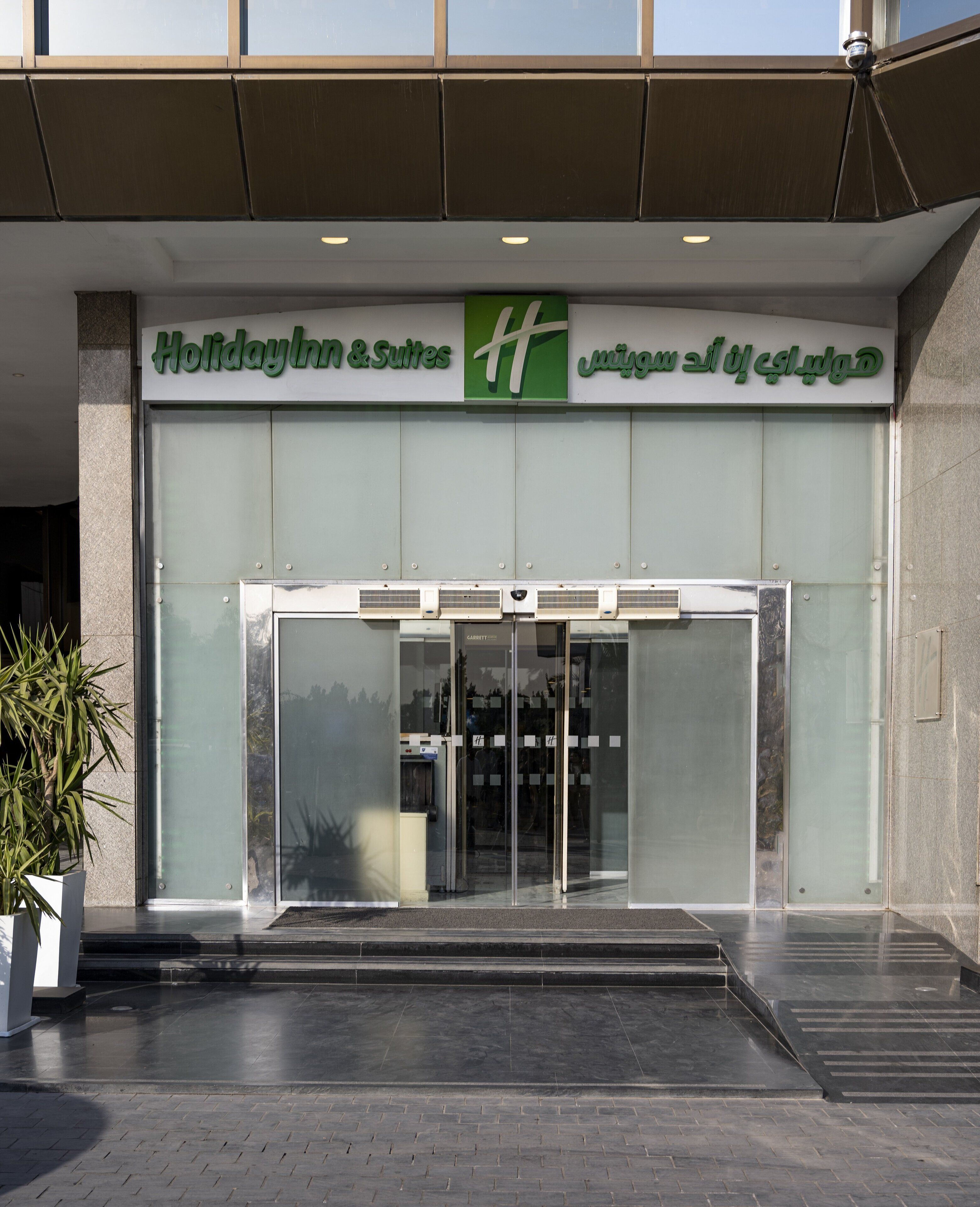 Entrada de hotel com porta de vidro, placa com a inscrição "Holiday Inn & Suites" e um logotipo verde.