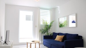 Uma sala de estar moderna com um sofá azul, uma mesa de madeira pequena, uma planta em vaso e uma janela grande com persianas.