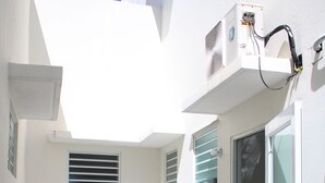 Um quarto claro, com paredes brancas, chão de azulejos, uma cadeira de madeira, uma mesinha e uma porta que dá para outro espaço.
