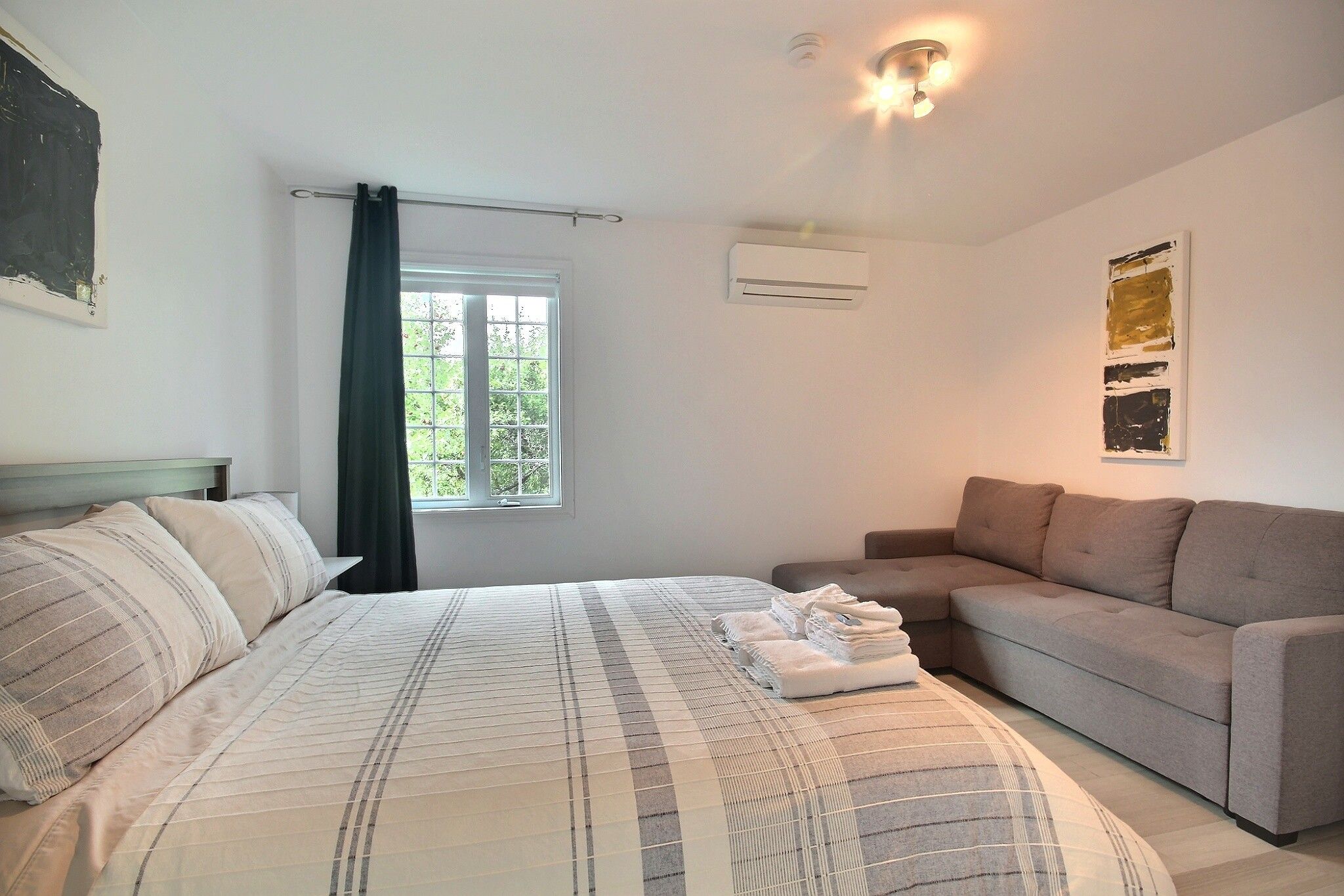 Quarto com cama, sofá cinza, janela com cortinas, ar condicionado e uma obra de arte pendurada na parede.