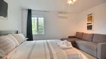Quarto com cama, sofá cinza, janela com cortinas, ar condicionado e uma obra de arte pendurada na parede.