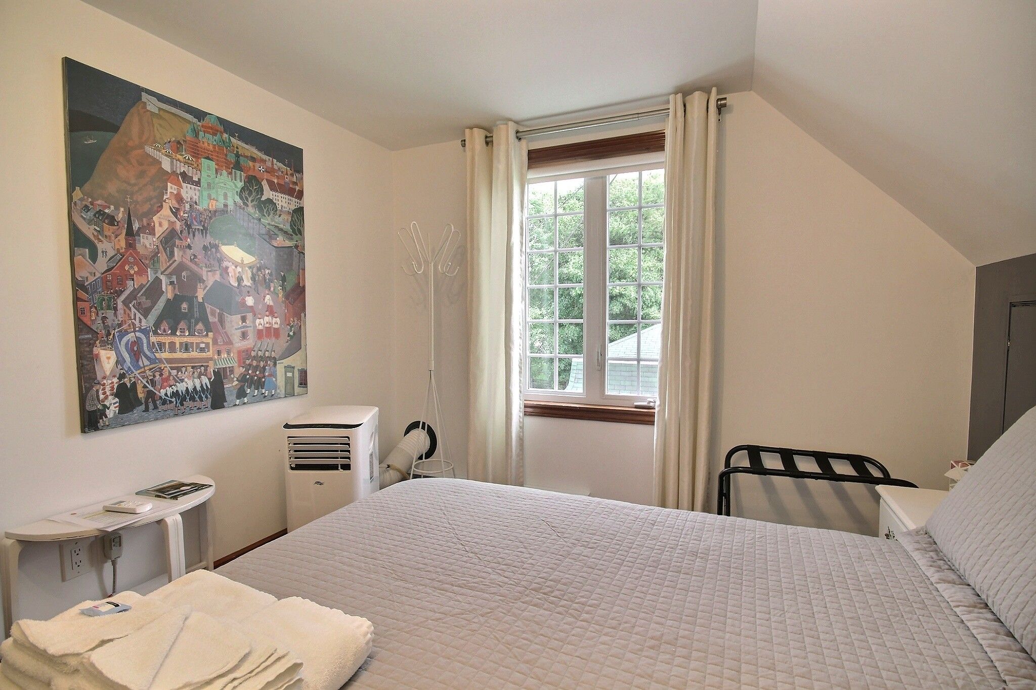 Quarto com uma cama grande, uma mesa de cabeceira, um radiador, uma janela com cortinas e um quadro na parede.