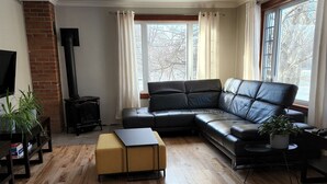 Uma sala de estar com piso de madeira, um sofá de couro preto, um banquinho amarelo, uma TV, uma lareira e uma janela grande com cortinas.