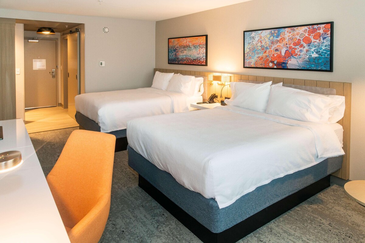 Quarto de hotel com duas camas, uma escrivaninha e uma cadeira. Há duas pinturas na parede e uma porta que dá para outro quarto.