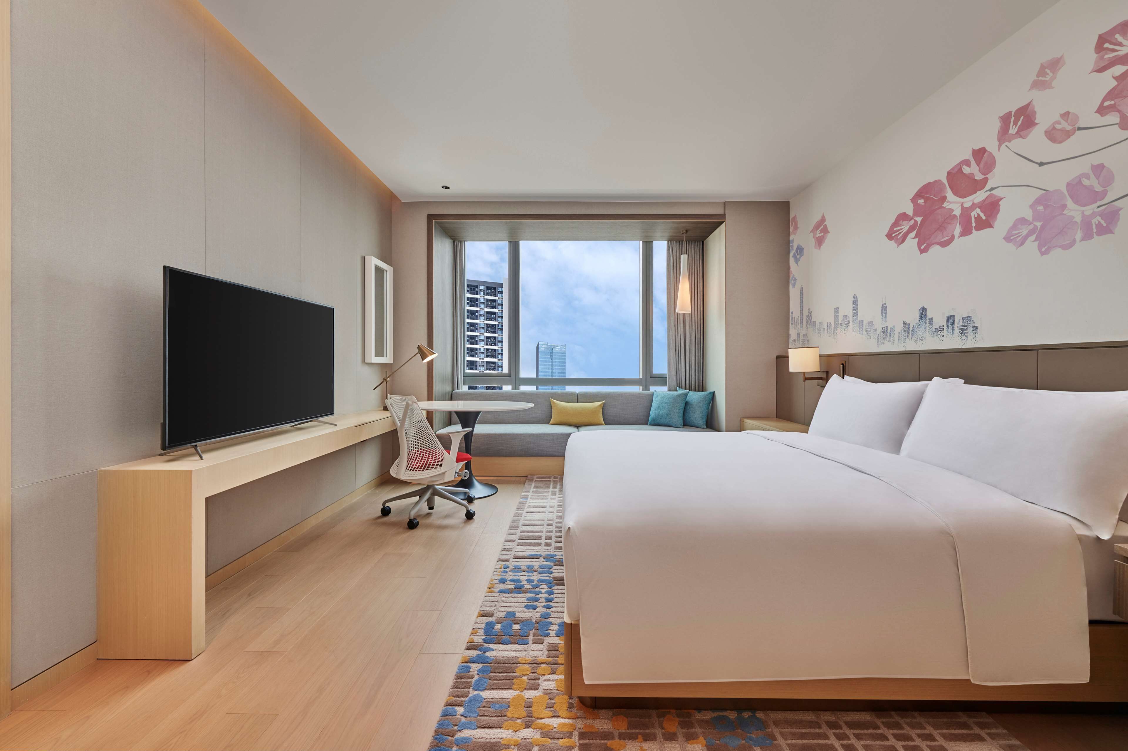 Quarto de hotel moderno com uma cama grande, televisão de tela plana, escrivaninha com cadeira e janela com vista para a cidade.