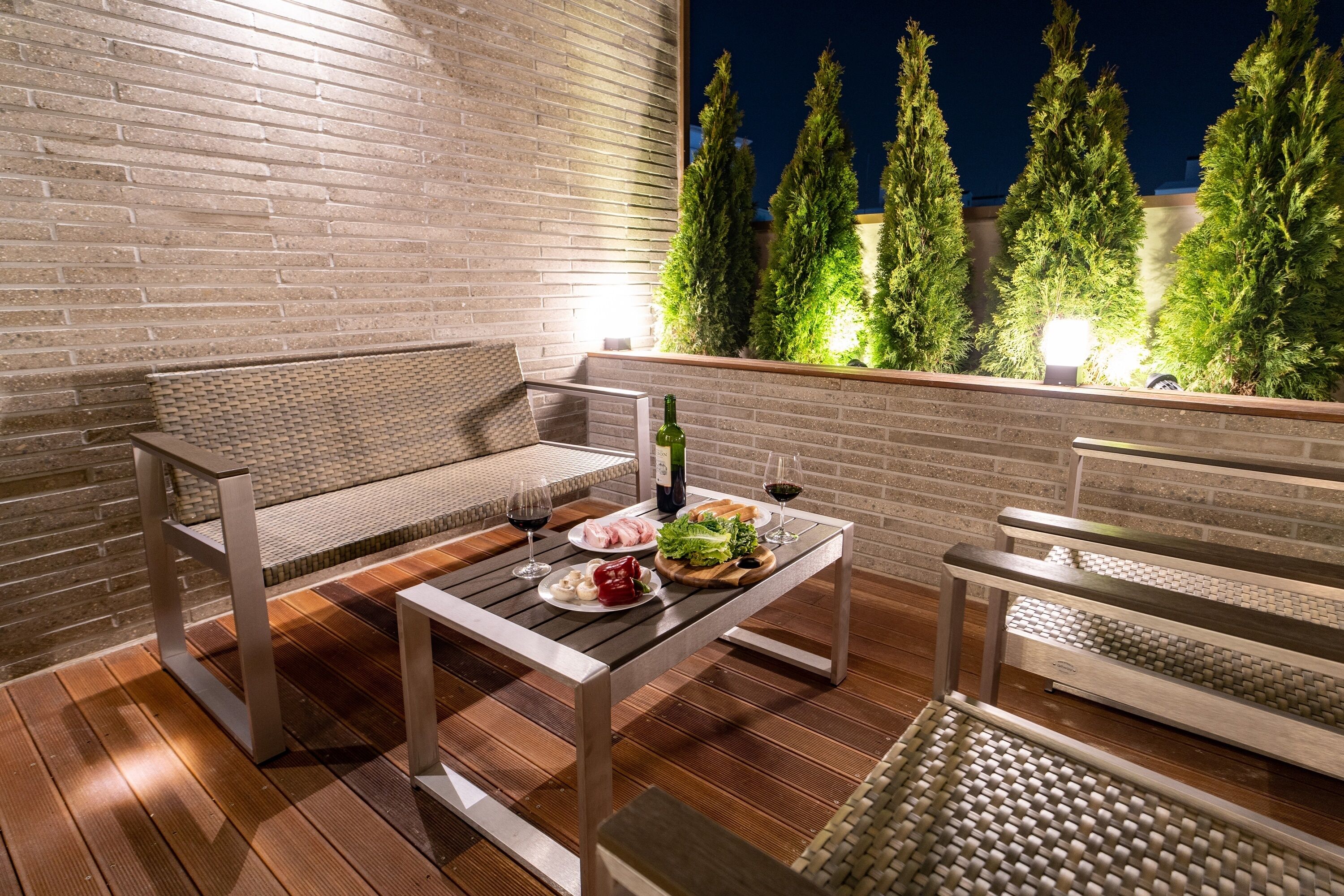 Um deck de madeira com sofá, uma mesa posta com comida e bebidas, e vista para árvores à noite.
