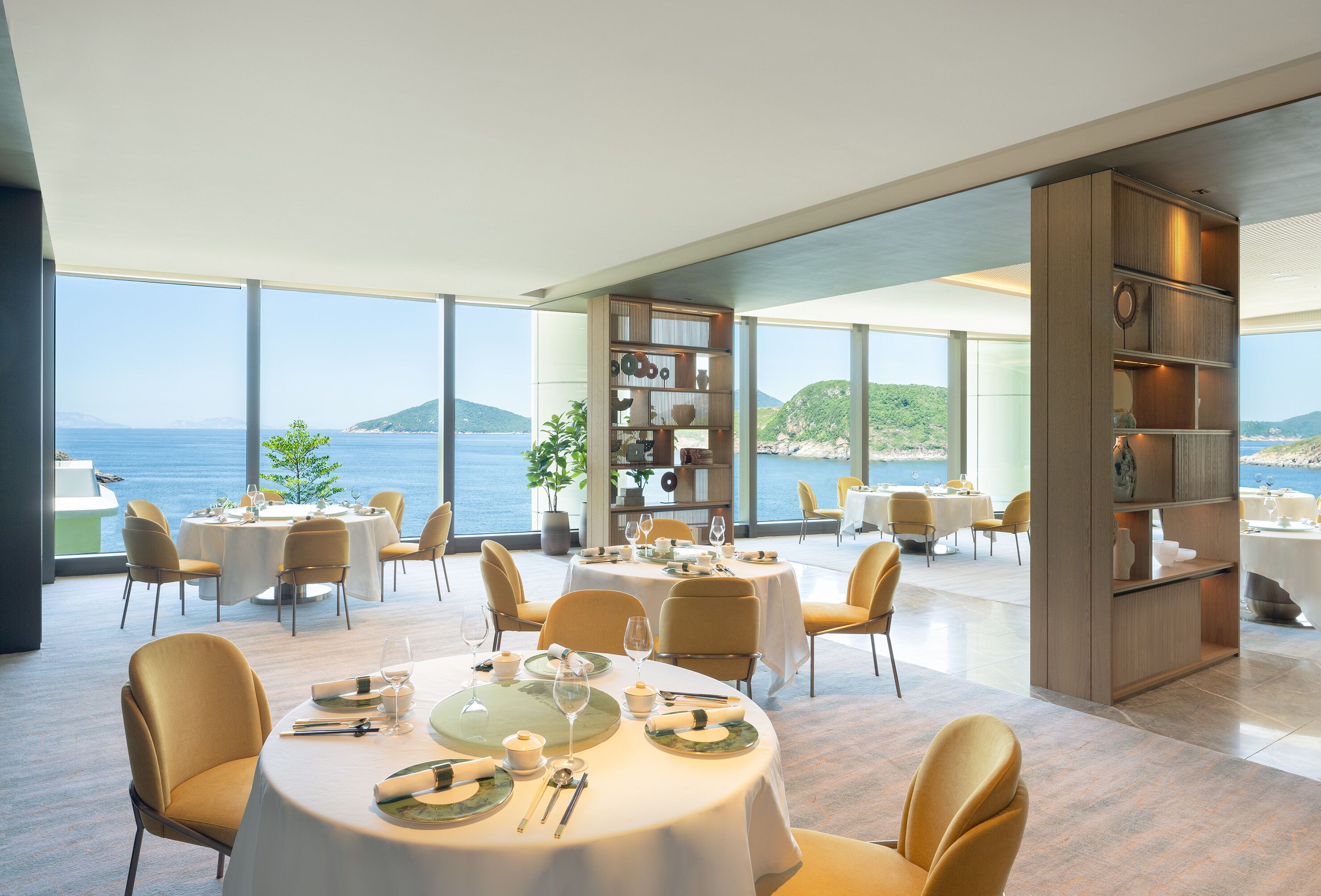 Um restaurante com mesas e cadeiras redondas, uma grande estante de livros e vista para o mar.