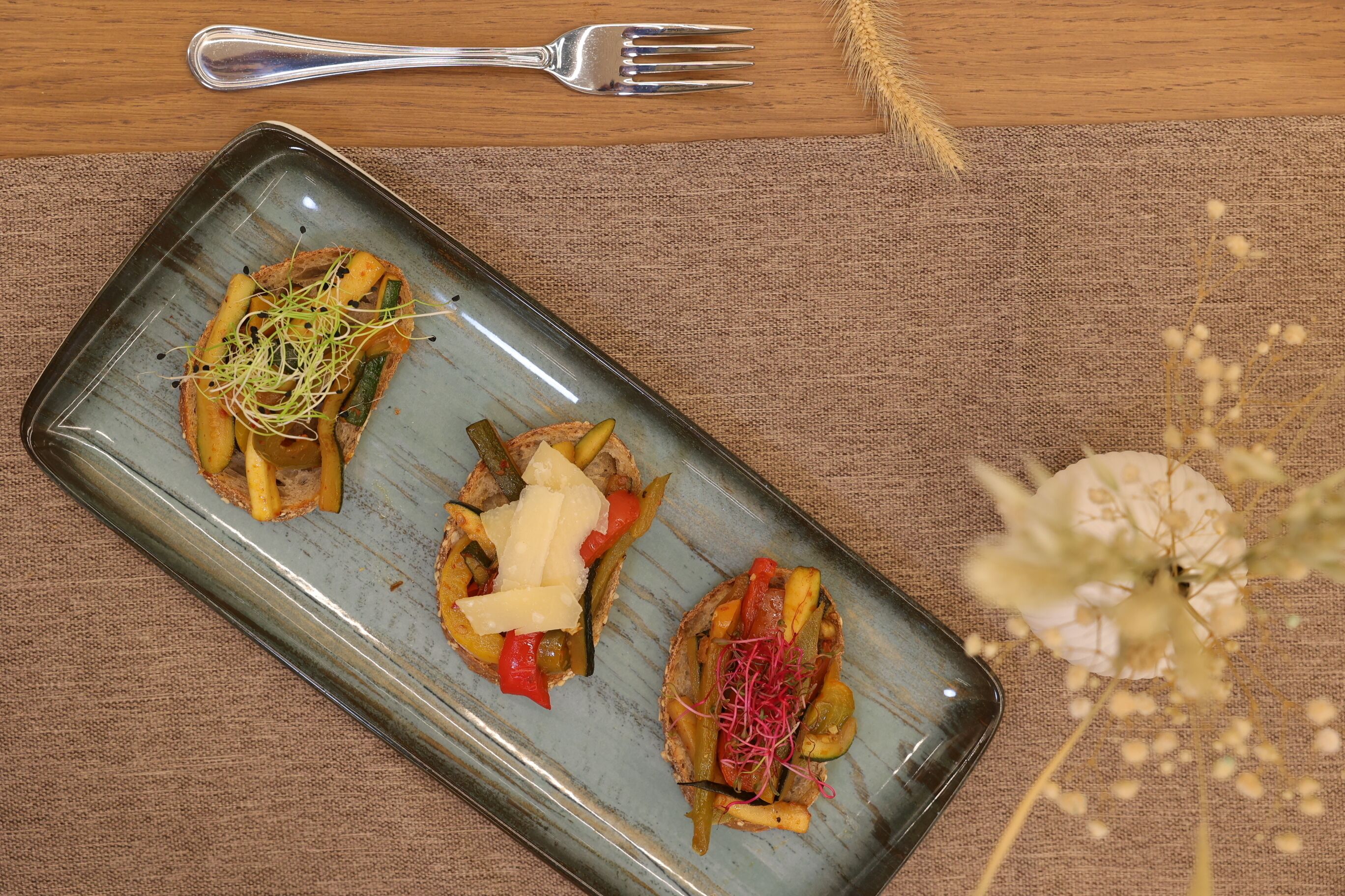 Prato retangular com três tipos diferentes de comida, um garfo e um vaso com flores secas sobre uma superfície texturizada.