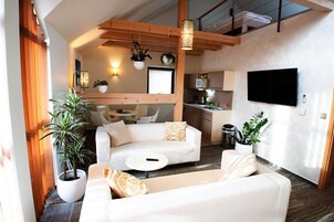 Sala de estar moderna com sofá branco, mesa de centro e TV fixada na parede. Plantas em vasos e uma área de cozinha são visíveis ao fundo.
