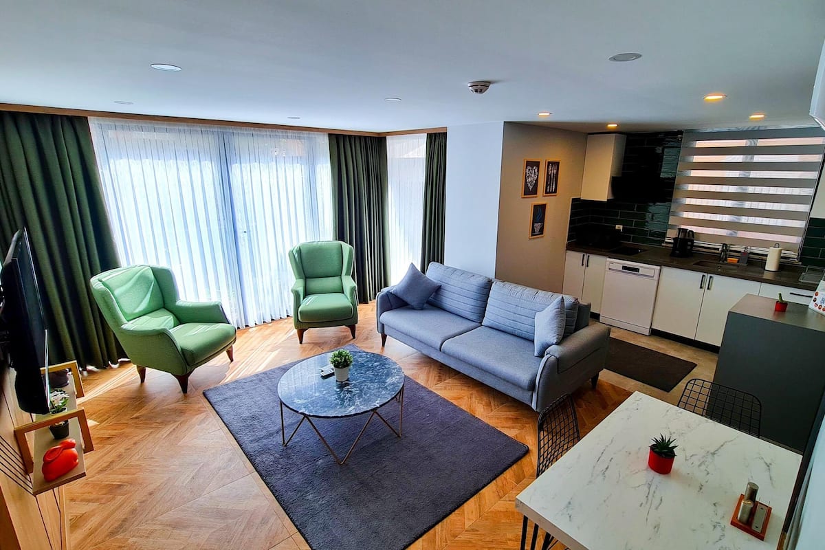 Uma sala de estar moderna com um sofá cinzento, duas poltronas verdes, uma mesa de centro e uma pequena área de cozinha com uma pia e fogão.