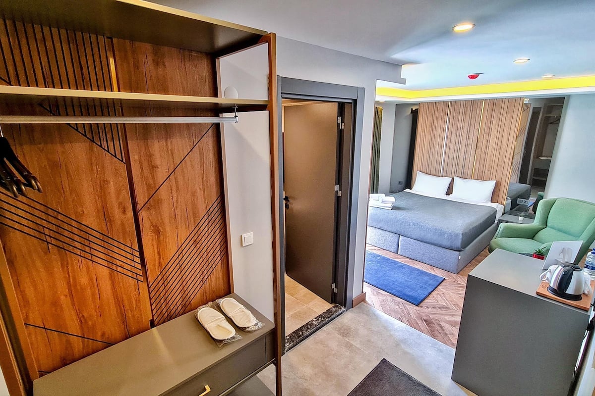 Quarto de hotel com cama, uma cadeira verde, escrivaninha com chaleira e guarda-roupa com roupas penduradas.