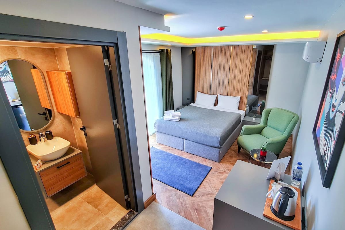 Quarto de hotel moderno com uma cama grande, uma poltrona verde, uma mesinha com chaleira e um banheiro com pia e espelho.